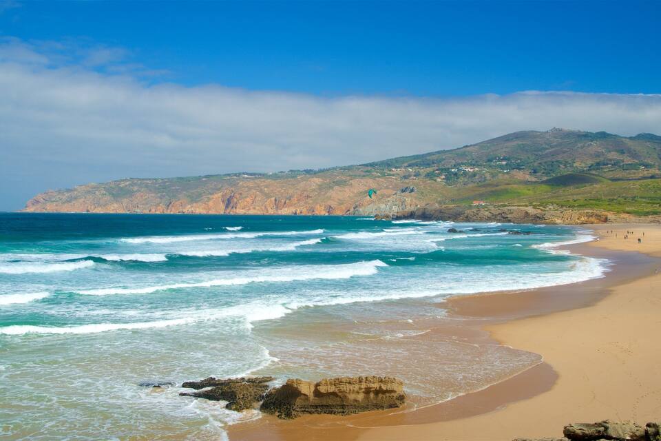 Uma praia arenosa com ondas, um afloramento rochoso e uma montanha ao fundo.