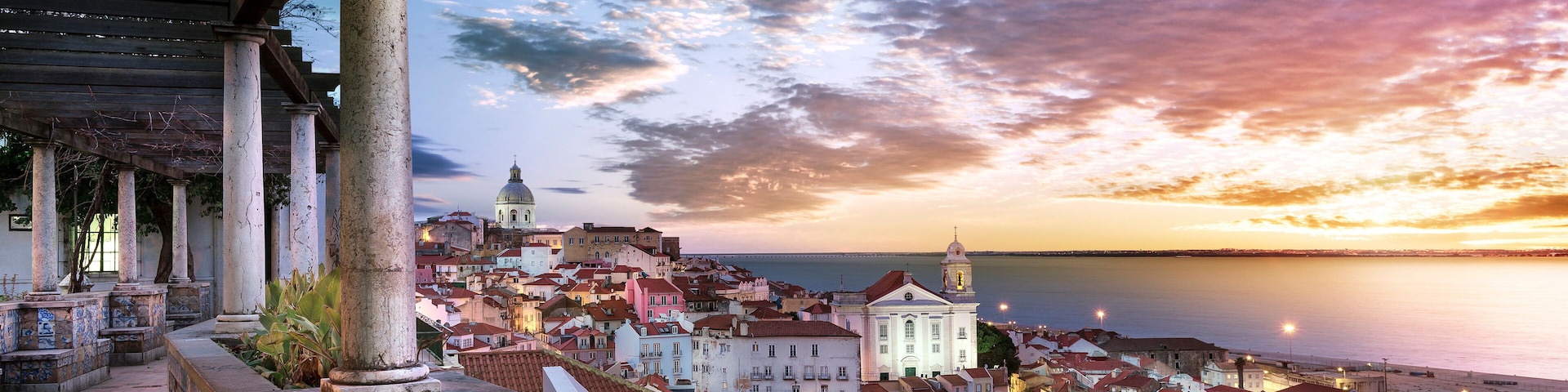 Paisagem urbana costeira ao pôr do sol, com um edifício abobadado em destaque e um calçadão de azulejos.