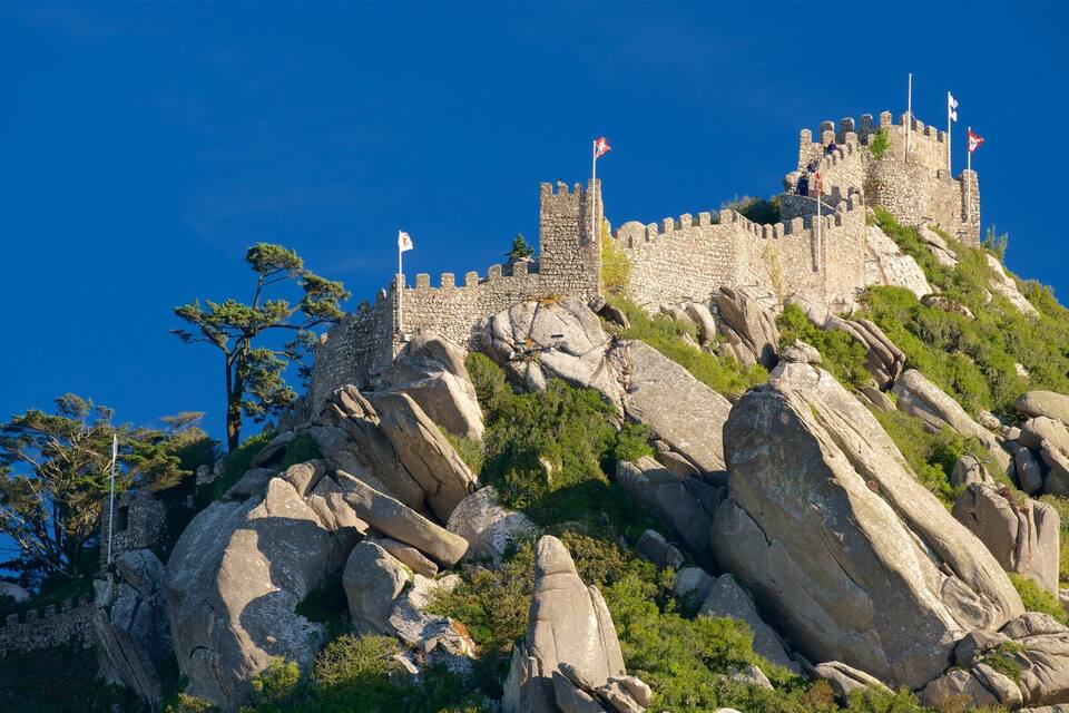 Um castelo de pedra histórico numa colina rochosa com bandeiras.