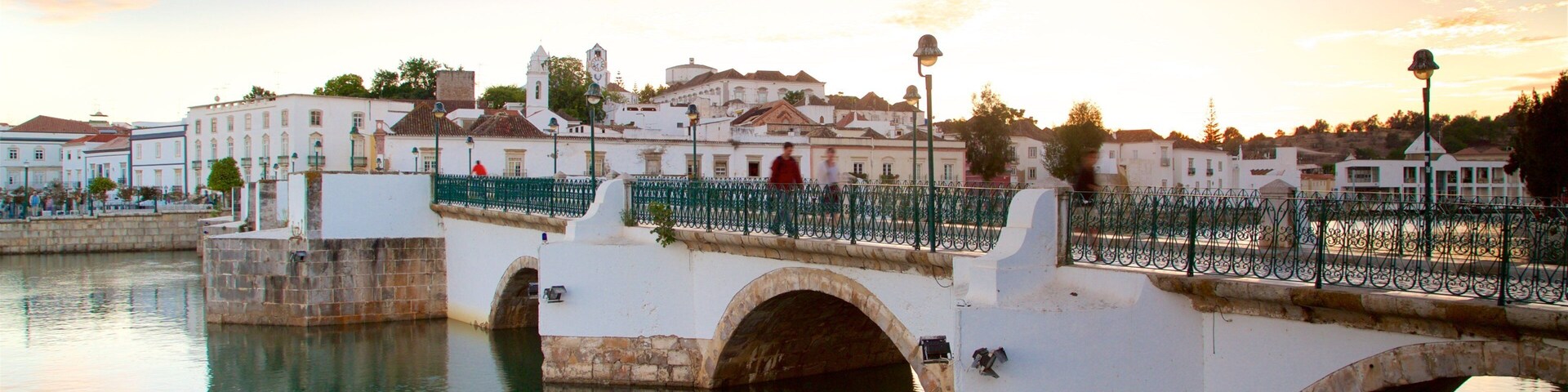Uma ponte de pedra sobre um rio calmo, com edifícios e um pôr do sol ao fundo.