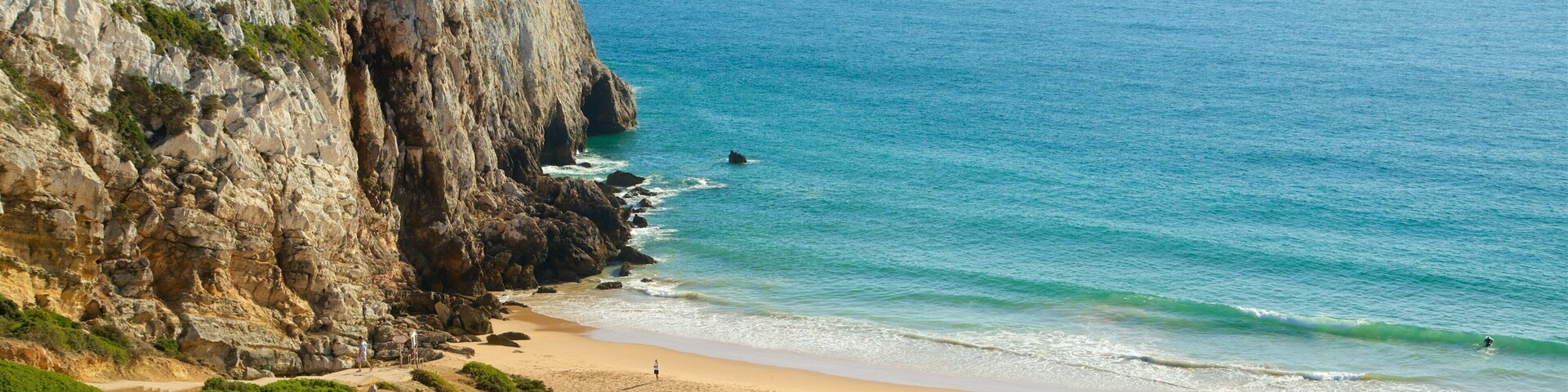 Paisagem costeira com uma praia arenosa, falésias rochosas e o oceano.