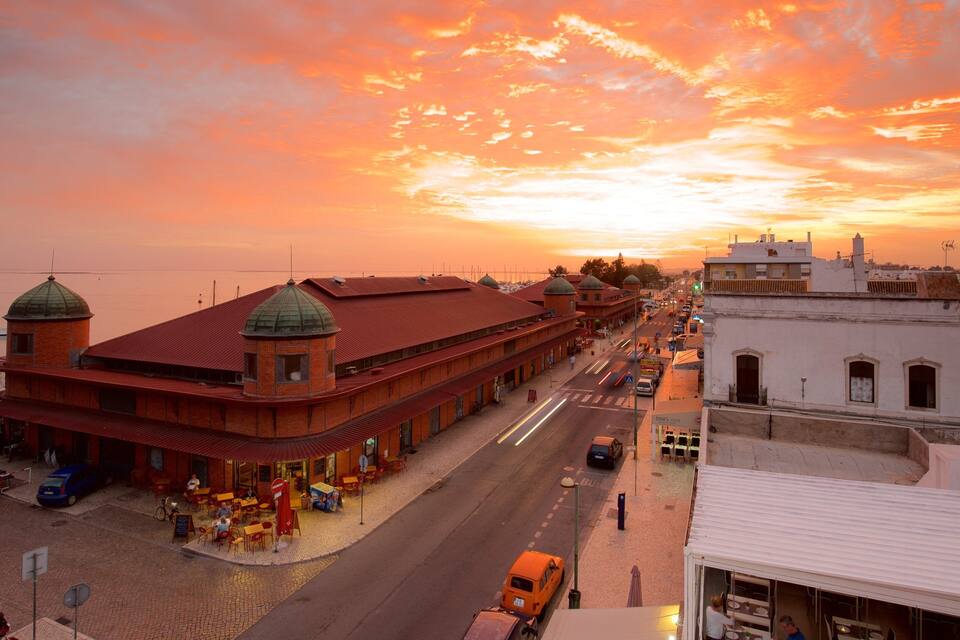 Rua à beira-mar ao pôr do sol, com prédios, um café e carros estacionados.