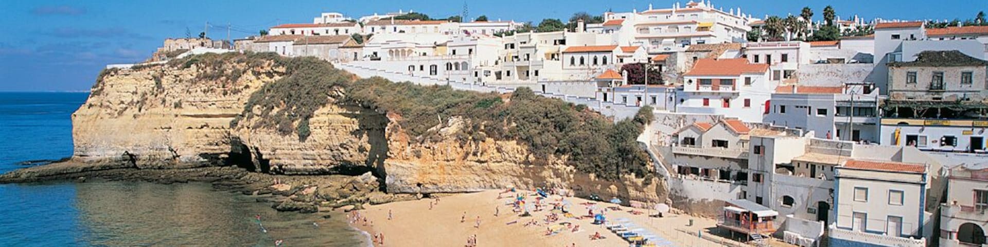 Uma vila costeira com uma praia arenosa, mar azul cristalino e edifícios brancos em um penhasco.