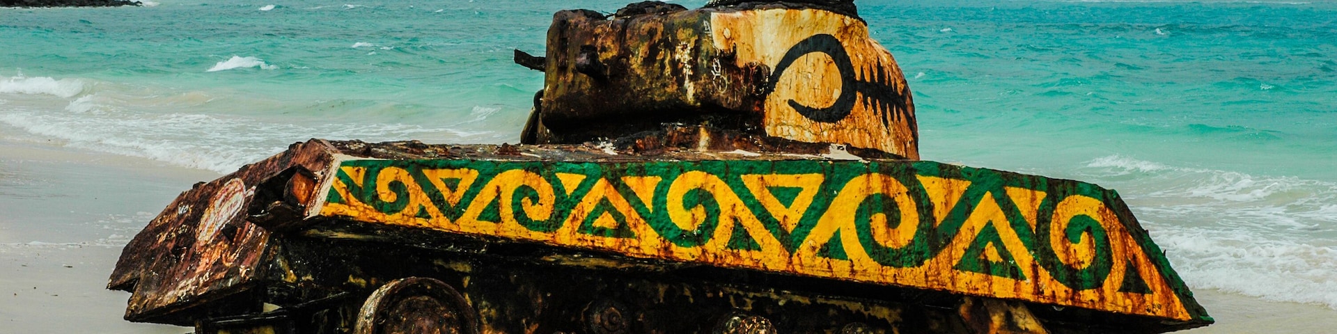 Um tanque militar enferrujado numa praia com água turquesa e céu nublado.