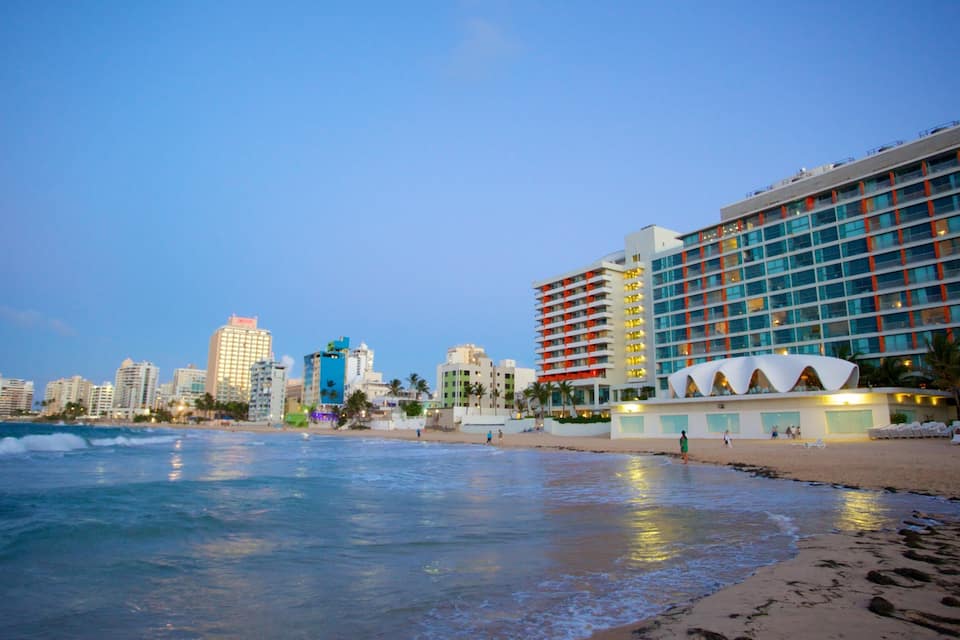 Uma praia com ondas do oceano, o horizonte de uma cidade e um hotel com fachada branca e laranja marcante.