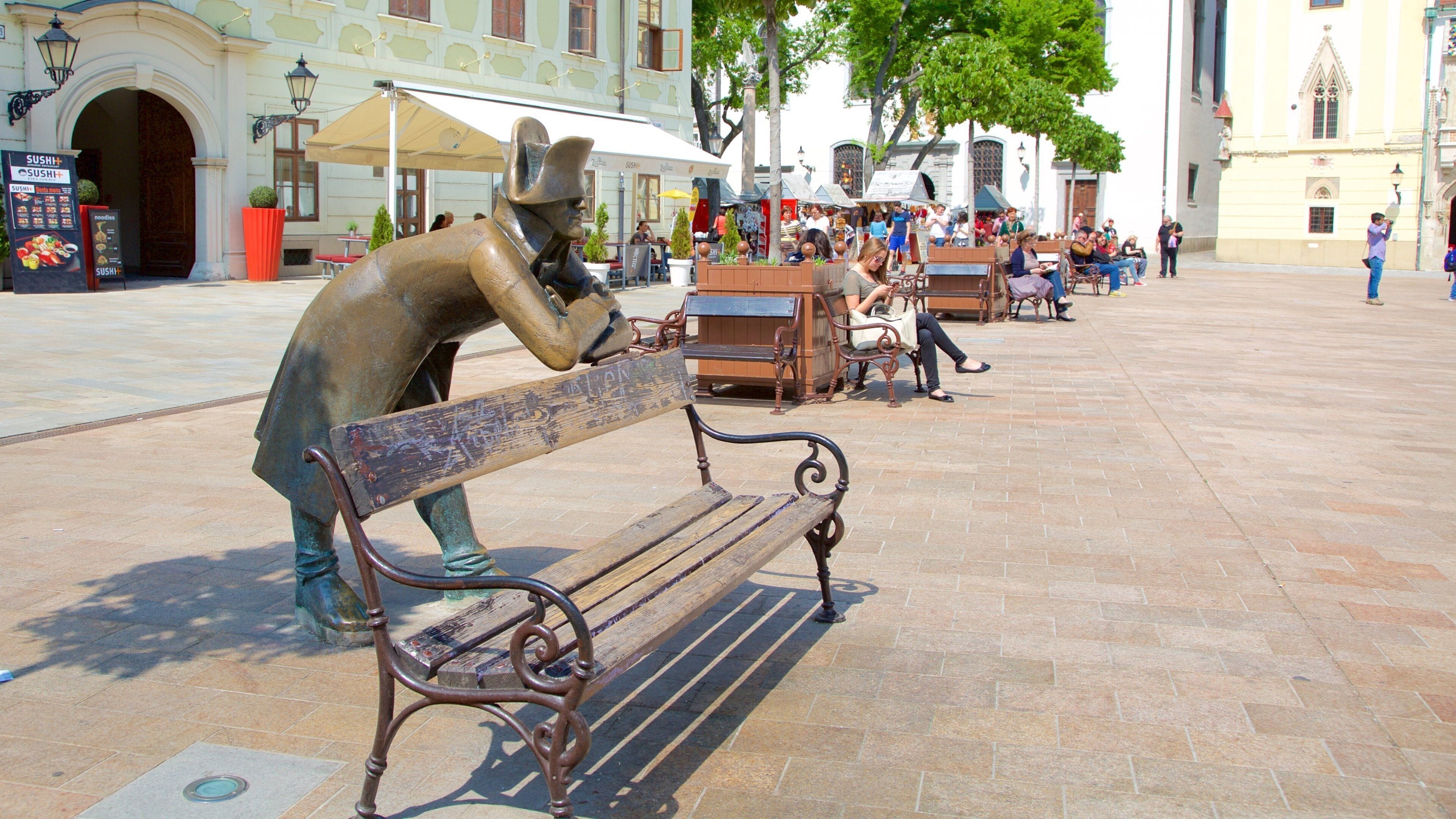 Estátua de bronze de um homem com chapéu sentado em um banco em praça pública, com pessoas ao redor.