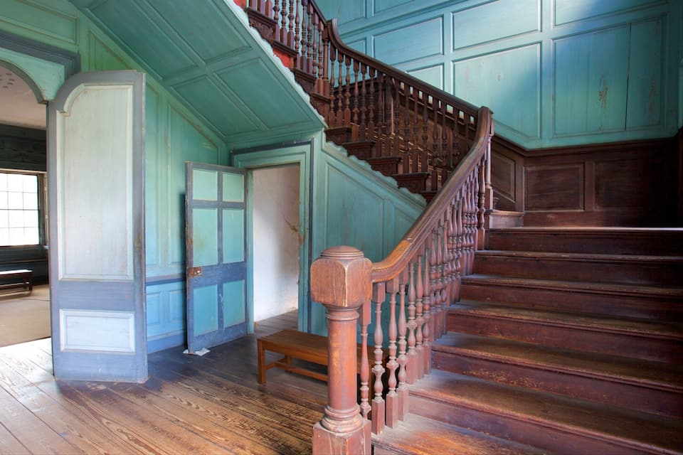 Uma escada de madeira com corrimão, um banco e uma porta em um quarto com paredes verde-água e piso de madeira.