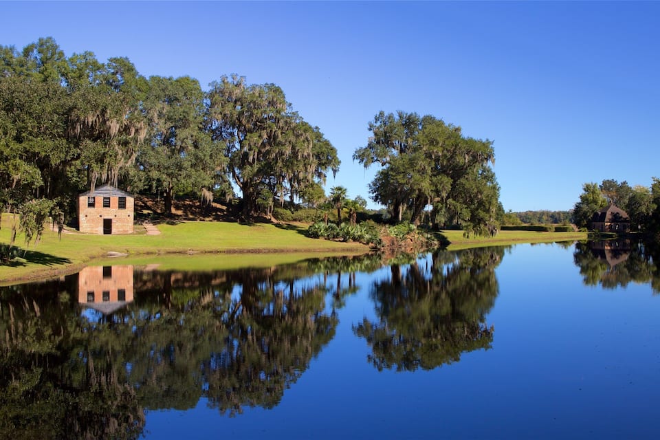 Um rio calmo com um pequeno edifício de tijolos e árvores exuberantes refletidas na água.