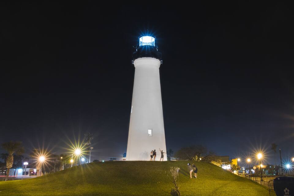 Um farol alto e branco, com luz azul no topo, rodeado por uma colina gramada e postes de luz.