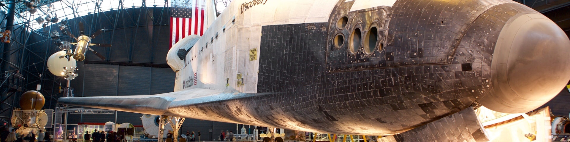 Um ônibus espacial em um museu, com uma grande bandeira americana e a palavra 'Discovery' na lateral.