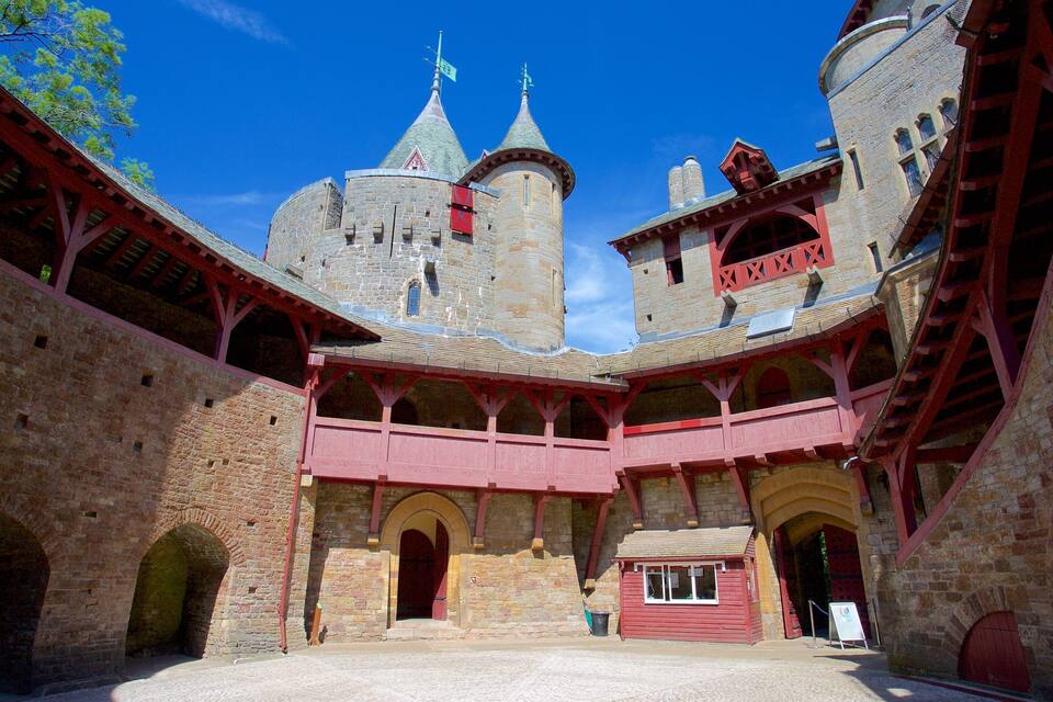 Pátio de castelo medieval com paredes de pedra, vigas de madeira vermelhas e uma torre central com telhados cônicos.
