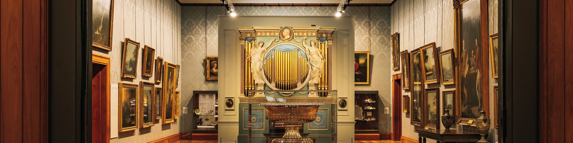 Uma galeria de arte com uma vitrine central ricamente decorada, ladeada por obras de arte emolduradas nas paredes.