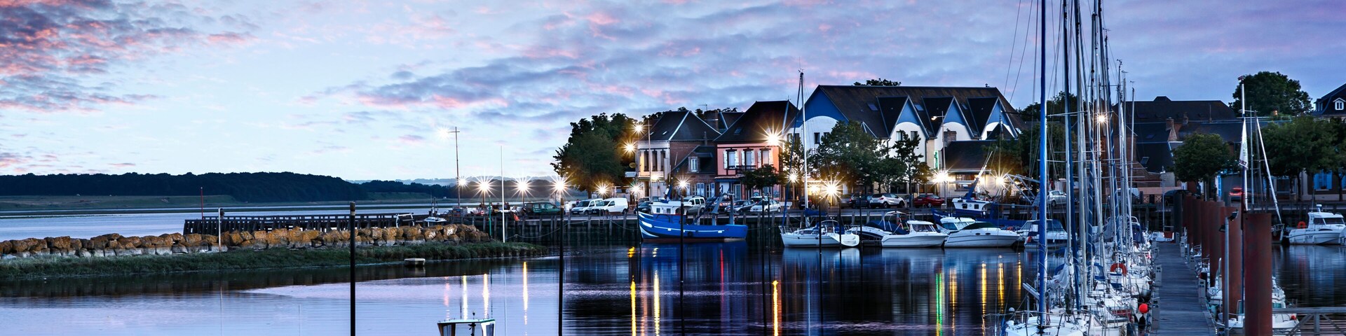 Um porto com barcos atracados, um cais e um edifício iluminado sob o céu crepuscular.
