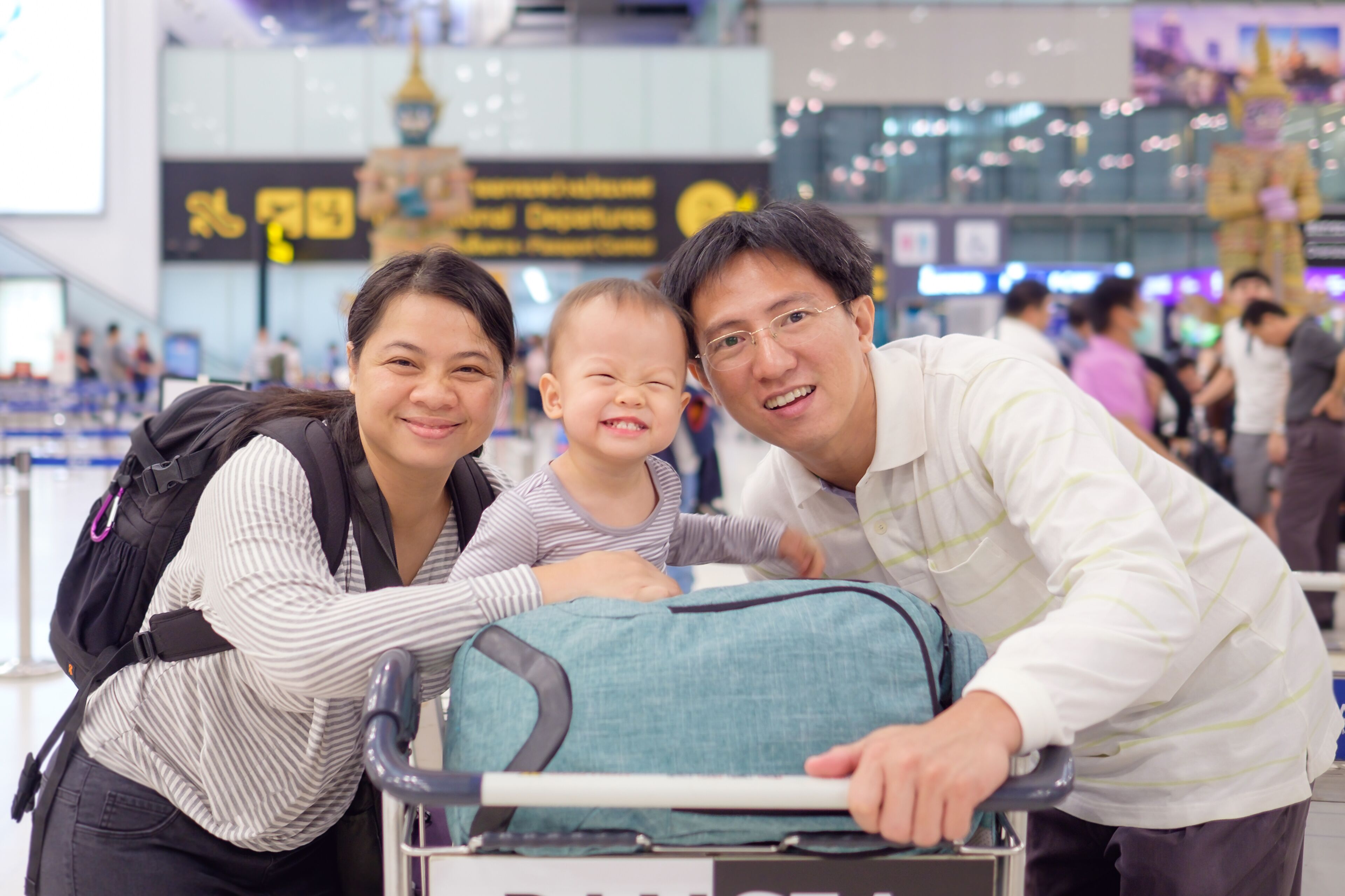 Uma família em um aeroporto com malas, uma placa com texto amarelo e uma grande estátua de Buda ao fundo.