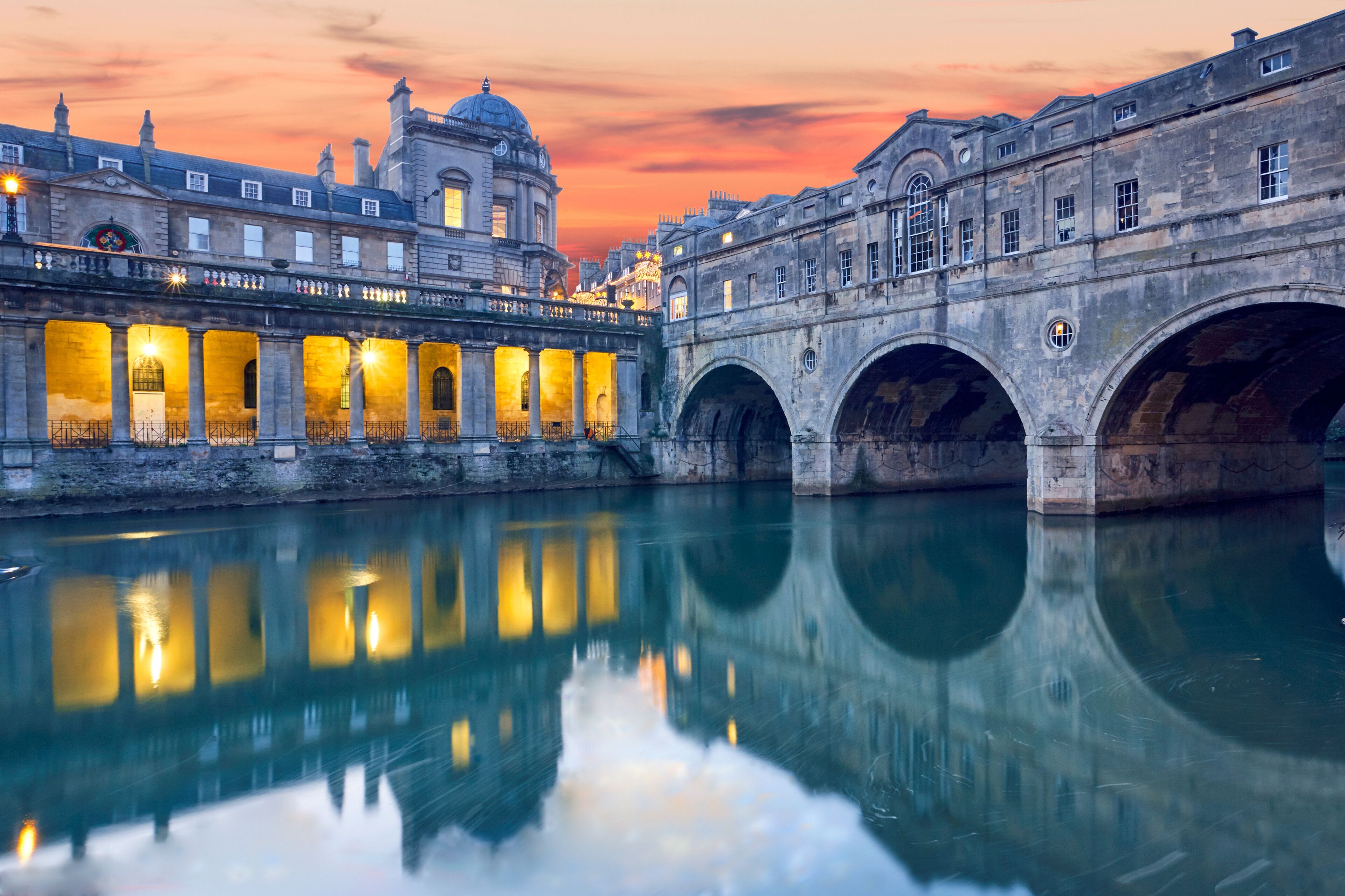 Uma ponte de pedra histórica sobre um rio calmo, refletindo edifícios e o céu.