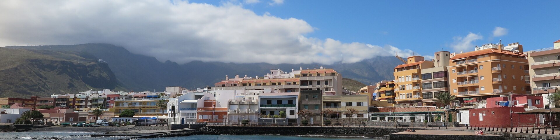 Uma cidade costeira com edifícios ao longo da orla, céu azul límpido e montanhas ao fundo.