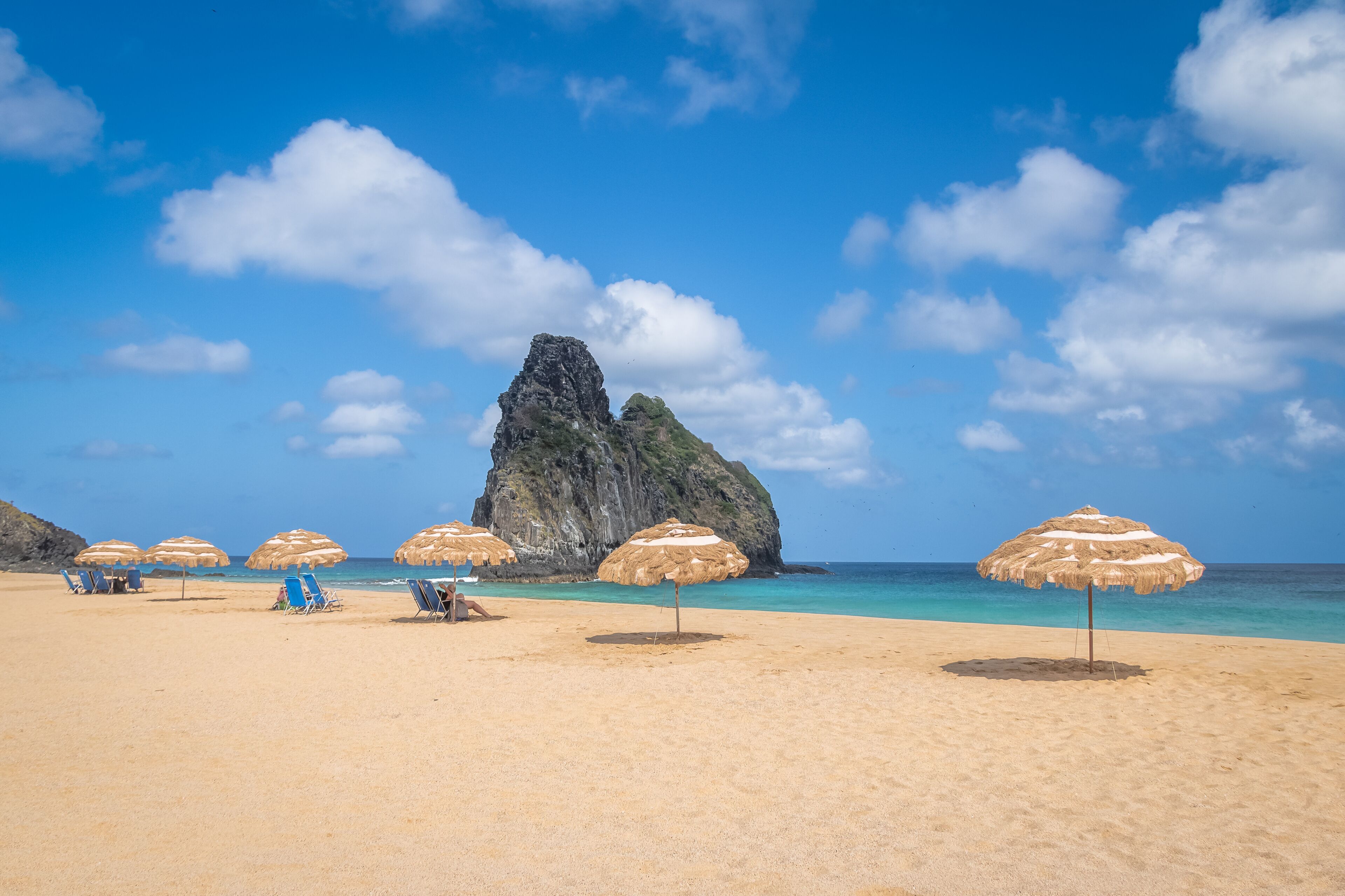 Uma praia com guarda-sóis de palha, águas turquesa cristalinas e uma rocha imponente.