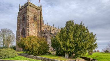 Uma igreja de pedra histórica com uma torre alta, cercada por vegetação bem cuidada e um cemitério.