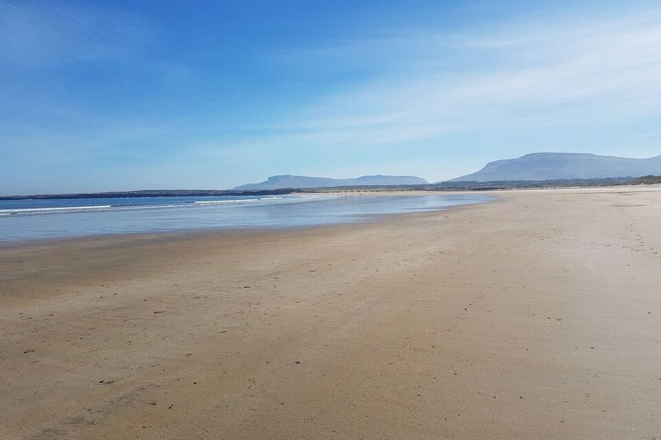 Uma extensa praia de areia com ondas suaves, montanhas ao longe e céu azul claro.
