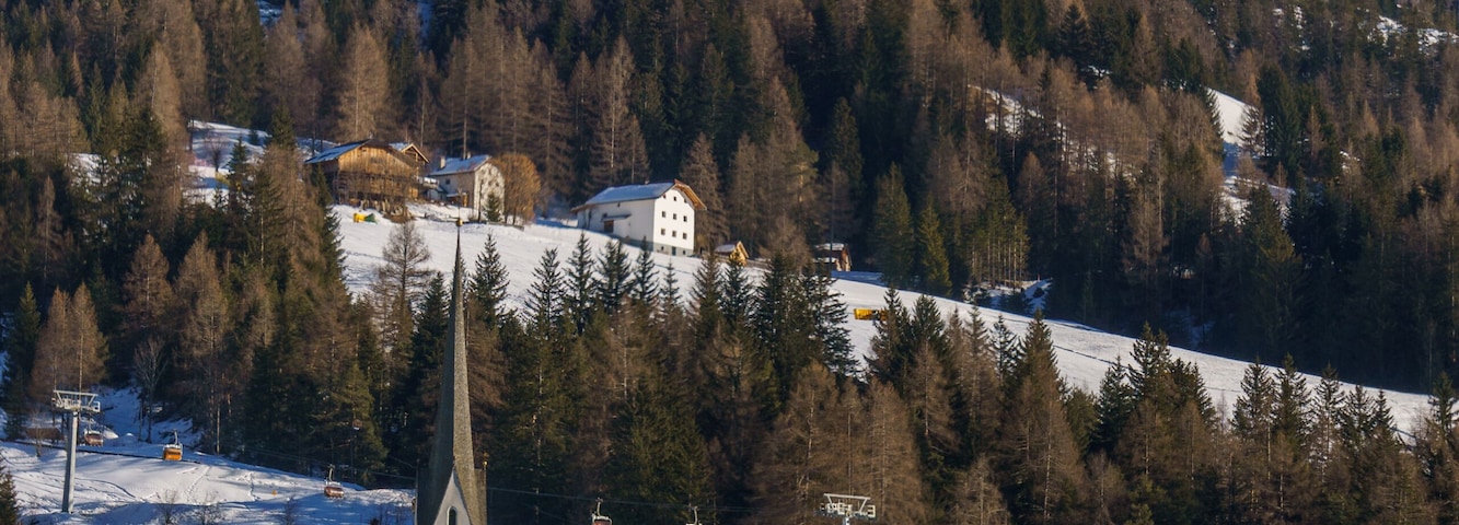 Vila coberta de neve nas montanhas, com uma igreja, casas e um teleférico de esqui, com montanhas acidentadas ao fundo.