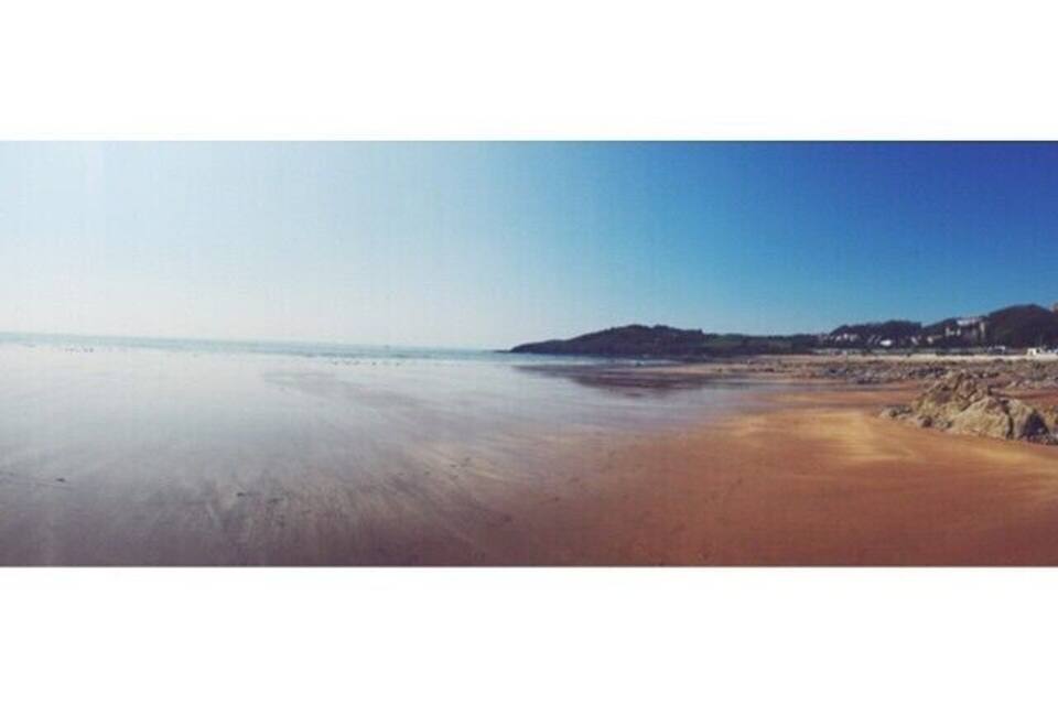 Uma praia com mar calmo, areal arenoso e colinas ao longe.