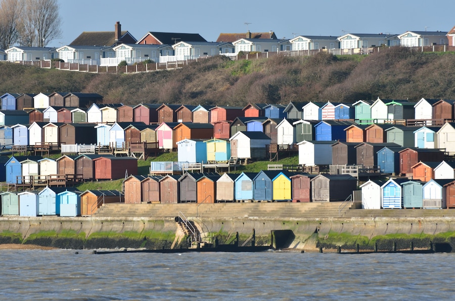 Uma fileira de cabanas coloridas à beira-mar, com casas ao fundo numa encosta.