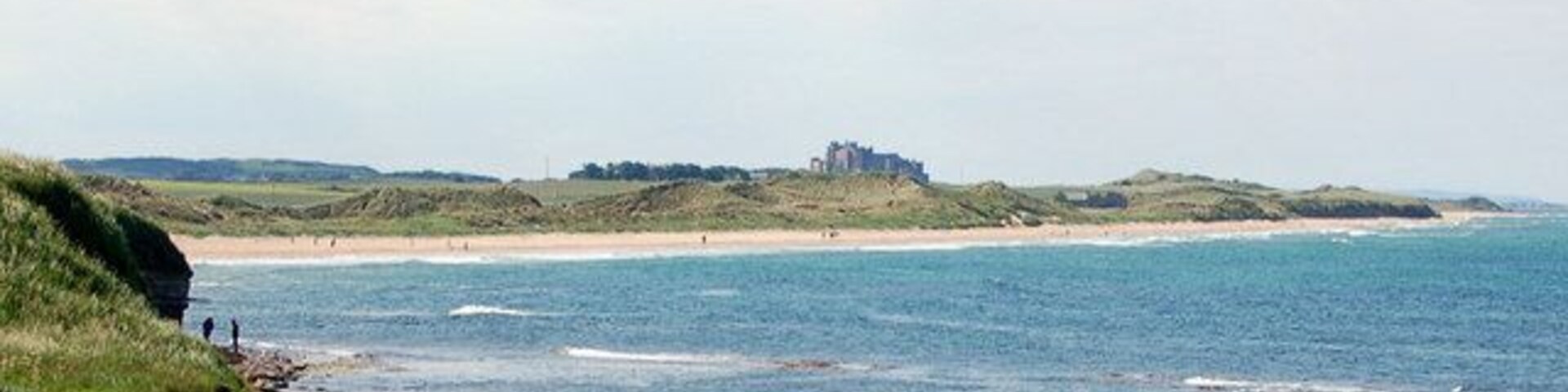 Uma praia rochosa com ondas, um edifício ao longe e colinas gramadas.
