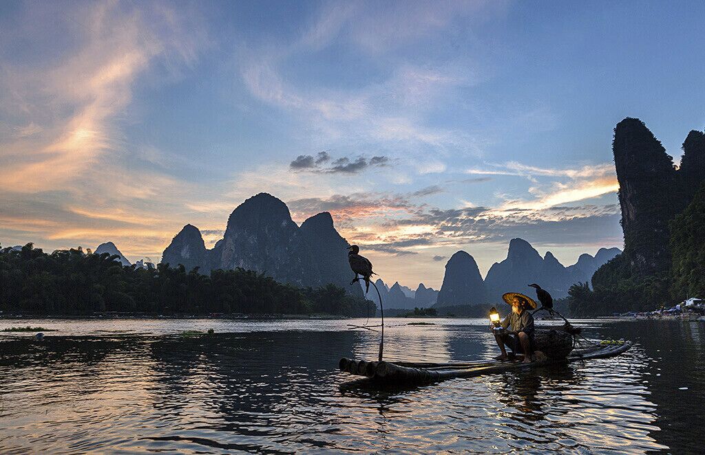 Um rio com um barco transportando duas pessoas e um pássaro, rodeado por falésias rochosas e um céu ao pôr do sol.