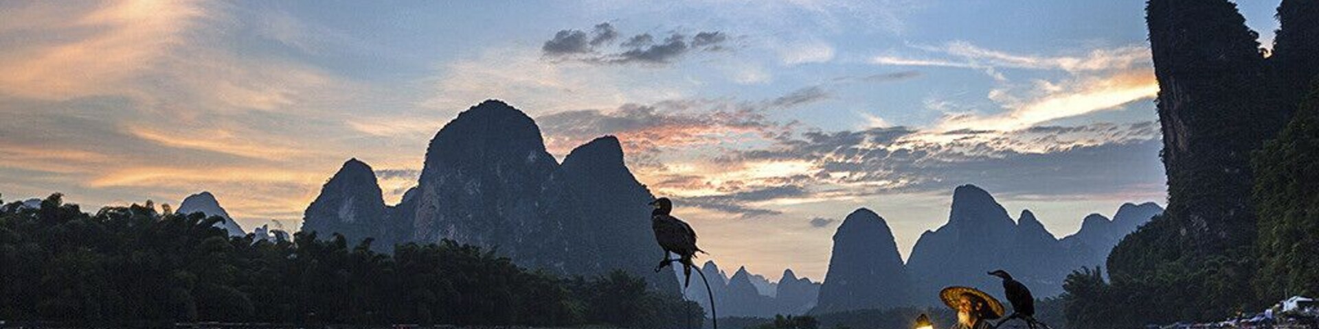 Um rio com um barco transportando duas pessoas e um pássaro, rodeado por falésias rochosas e um céu ao pôr do sol.