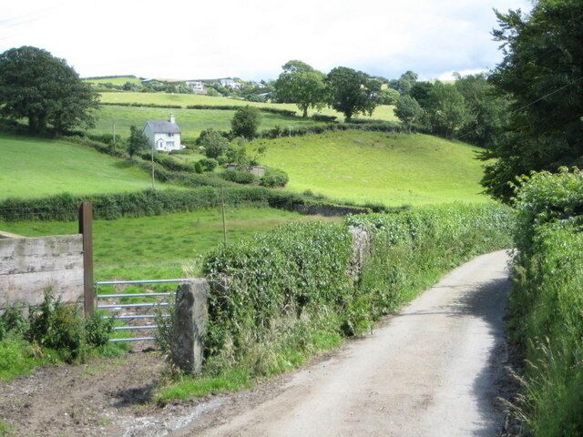 Uma estrada rural com um portão de pedra, campos verdes exuberantes e uma casa branca ao longe.