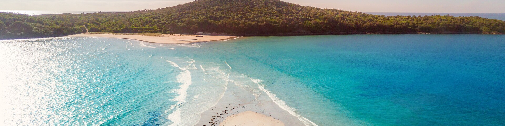 Paisagem costeira com praia arenosa, águas turquesa cristalinas e uma colina coberta de vegetação.