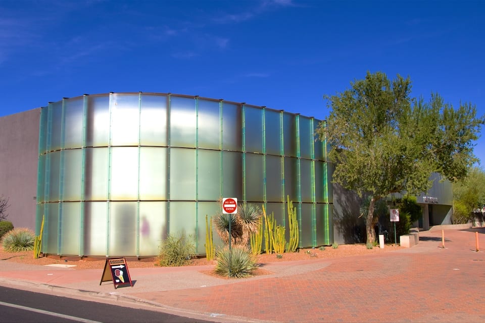 Um edifício moderno com superfície refletora, rodeado por vegetação desértica e um céu azul límpido.