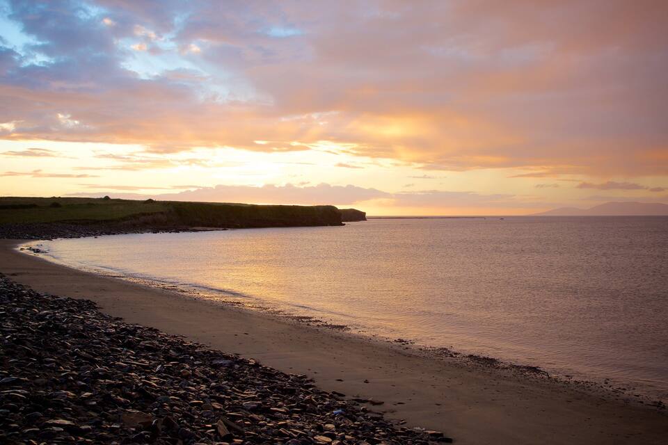 Uma praia de seixos com um litoral rochoso e um pôr do sol sobre a água.