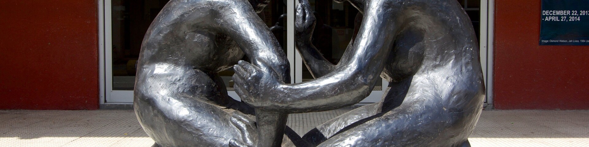 Escultura de duas figuras em pose contemplativa em frente a um edifício com a placa 'National Gallery of Jamaica'.