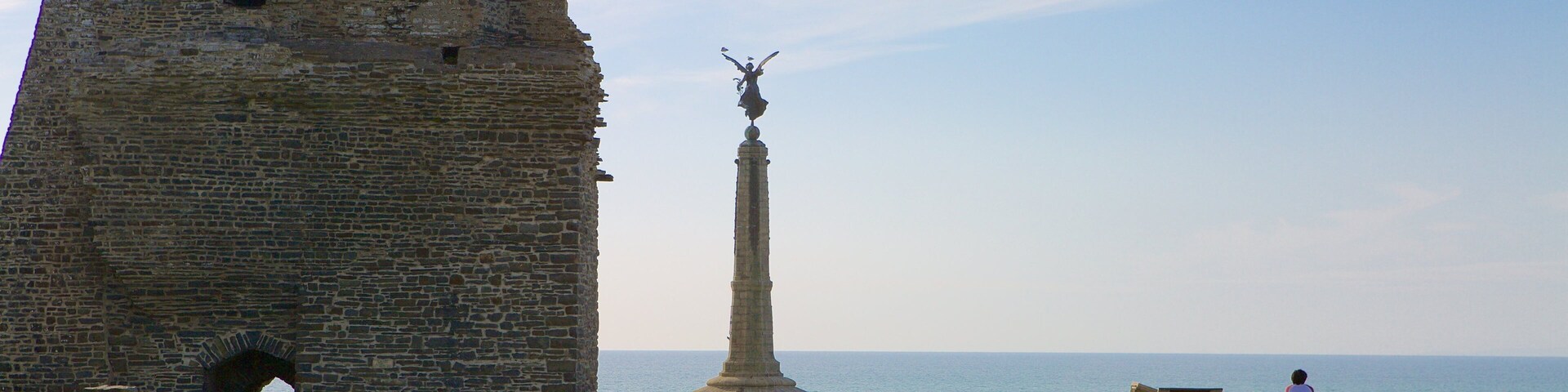 Uma torre de pedra, um monumento com uma estátua, e uma pessoa sentada na relva junto ao mar.