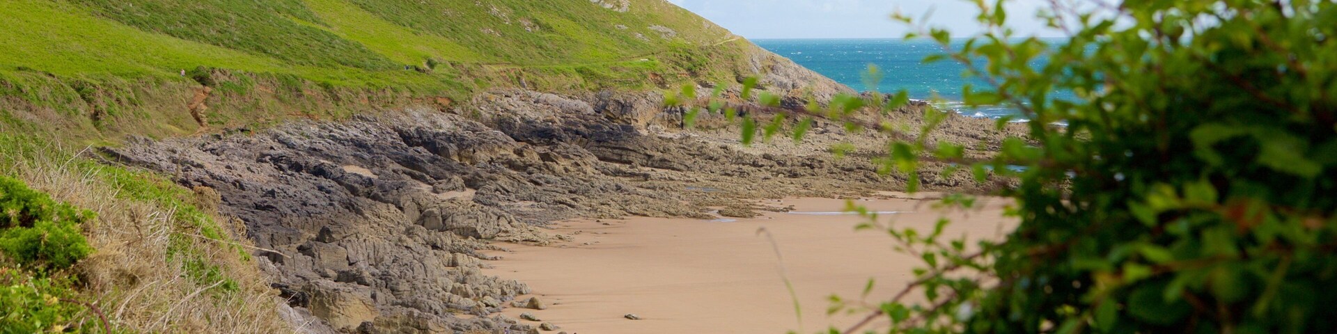 Paisagem costeira com uma praia de areia, falésias rochosas e colinas verdes.
