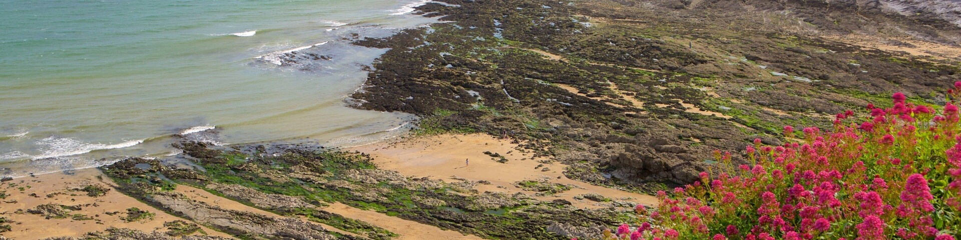 Paisagem costeira com costas rochosas, uma praia de areia e uma colina com um edifício no topo.