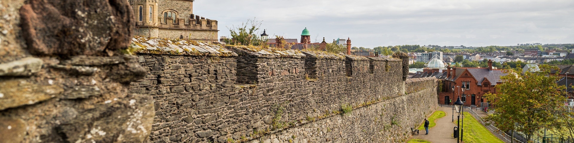 Um muro de pedra com uma estrutura semelhante a um castelo no topo, um caminho e uma vista distante de uma cidade.