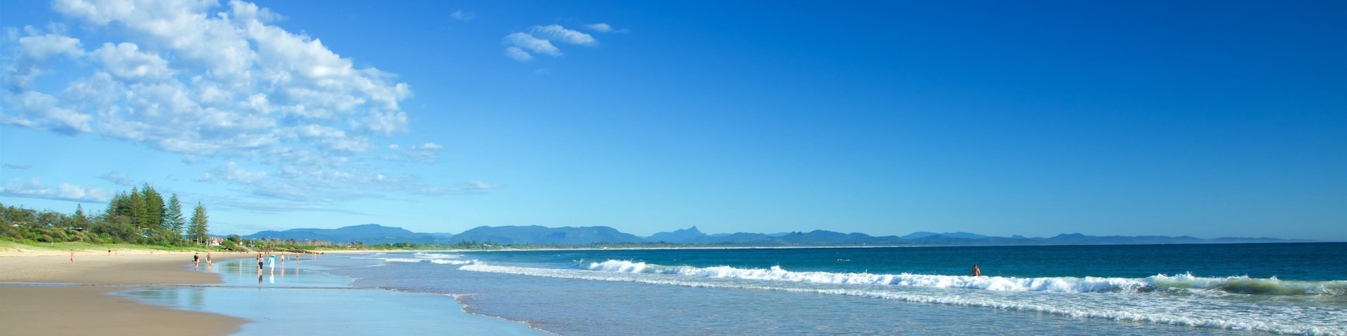 Uma praia com águas azul-claras, areal arenoso e montanhas ao longe sob um céu parcialmente nublado.