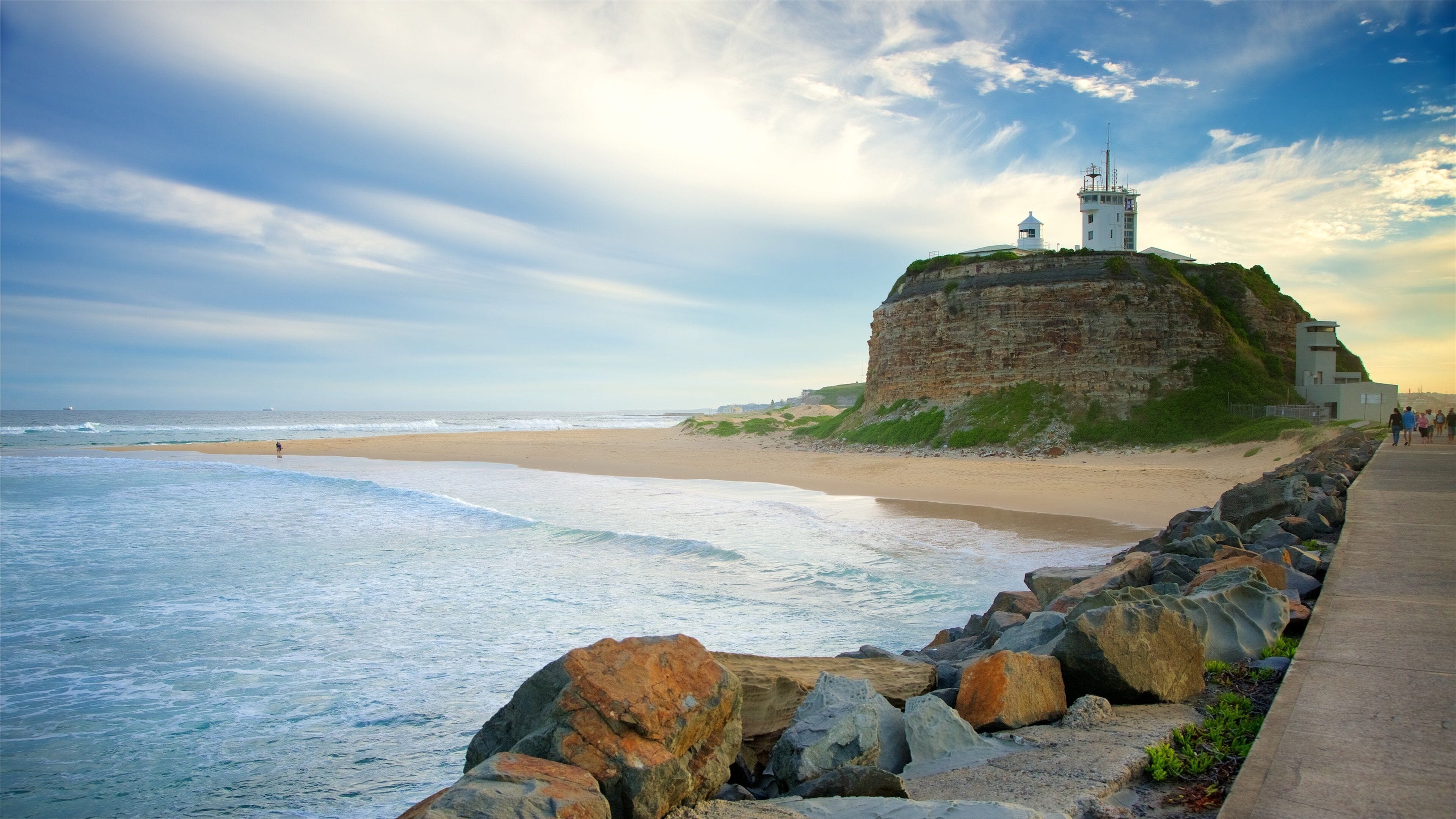 Cena costeira com um farol em um penhasco, uma praia de areia e um molhe rochoso.