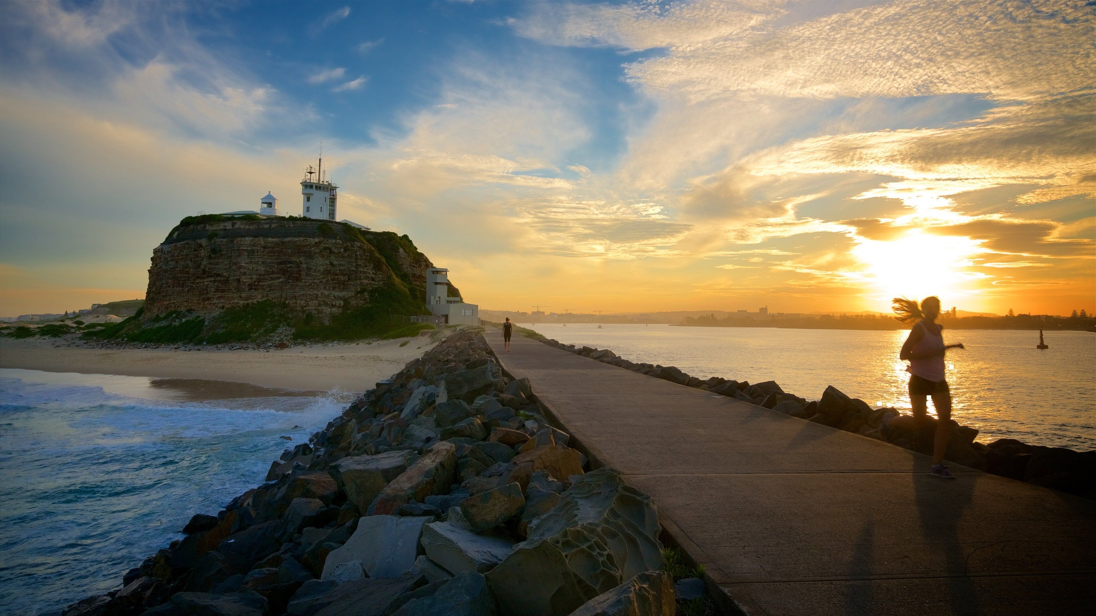 Um pôr do sol costeiro com um farol, margem rochosa e um calçadão pavimentado.