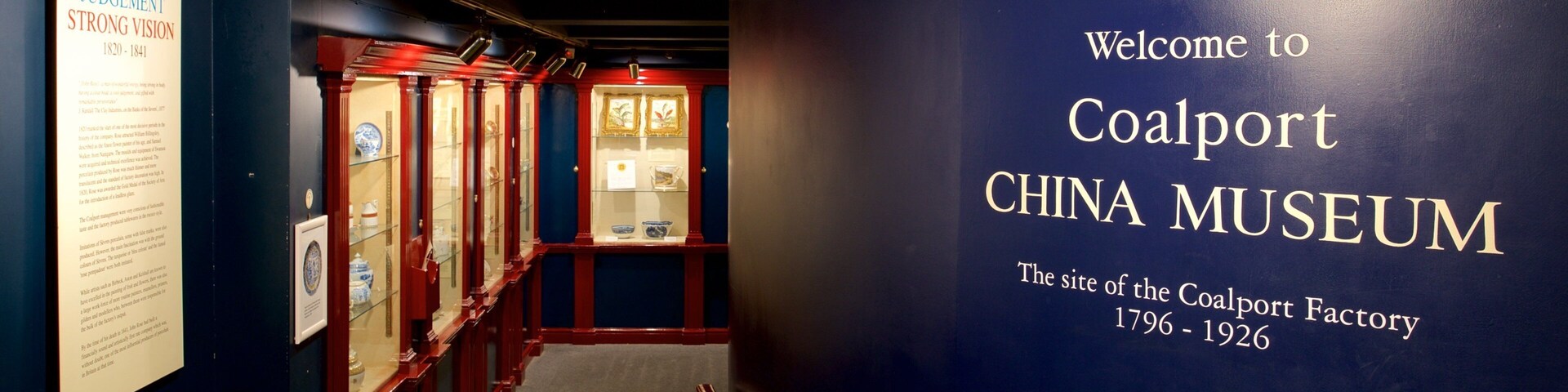 Corredor com vitrines e uma grande parede azul com o texto 'Welcome to Coalport China Museum'.