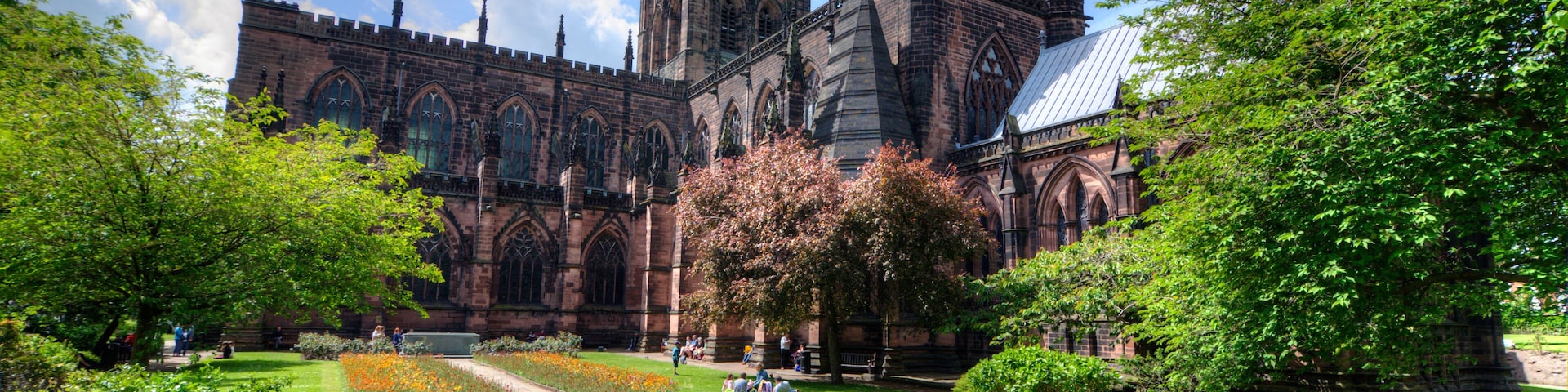 Uma catedral histórica com arquitetura elaborada, rodeada por um jardim bem cuidado com canteiros floridos e um padrão circular de pedra no centro.