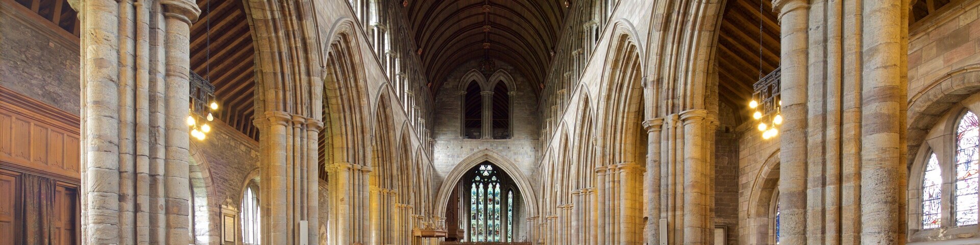 Interior de uma catedral com tetos abobadados altos, janelas em arco e fileiras de bancos de madeira.