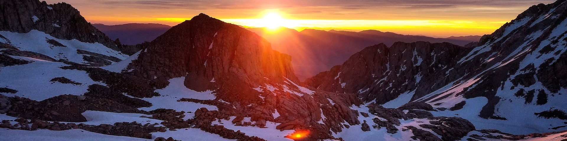 Paisagem montanhosa com um pôr do sol, picos cobertos de neve e um vale.