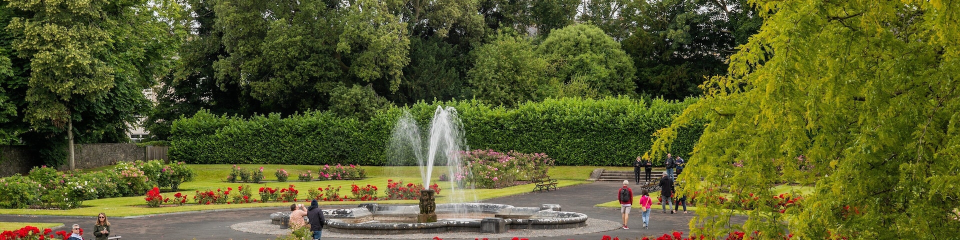 Um parque com uma fonte, flores vermelhas e pessoas caminhando.