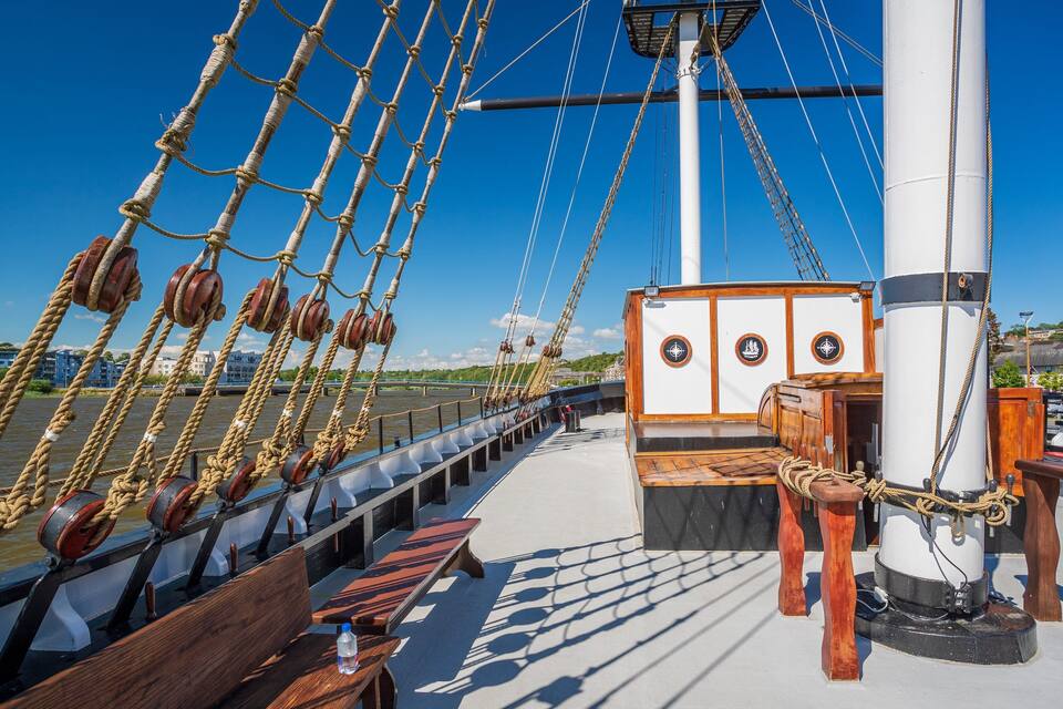 Convés de navio com bancos de madeira, cordas e um pilar branco.