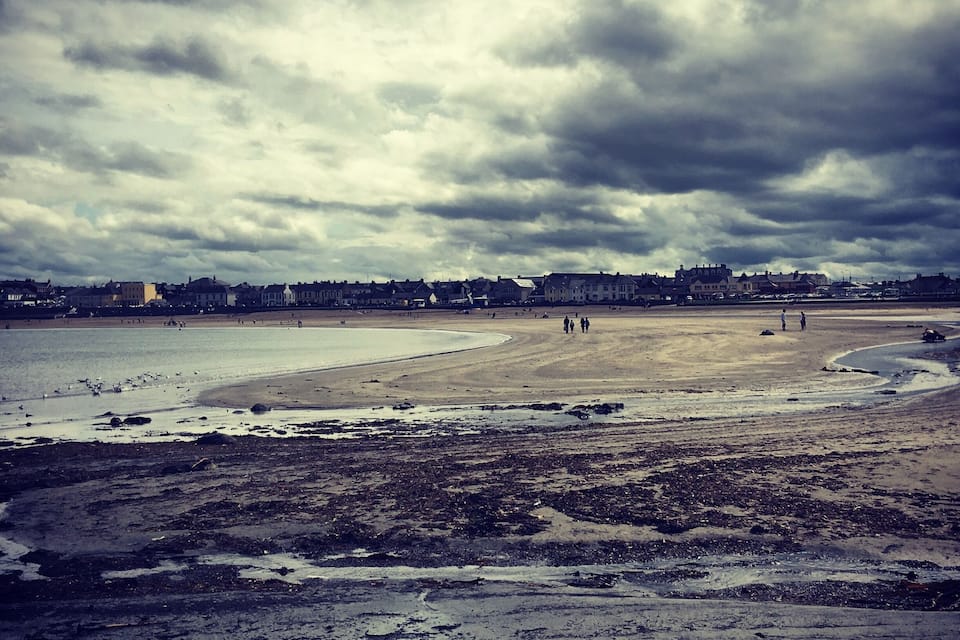 Uma praia com céu nublado, areal e edifícios ao fundo.