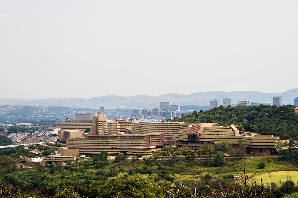 Um complexo amplo de edifícios com vários andares, rodeado por vegetação exuberante e colinas ao fundo.