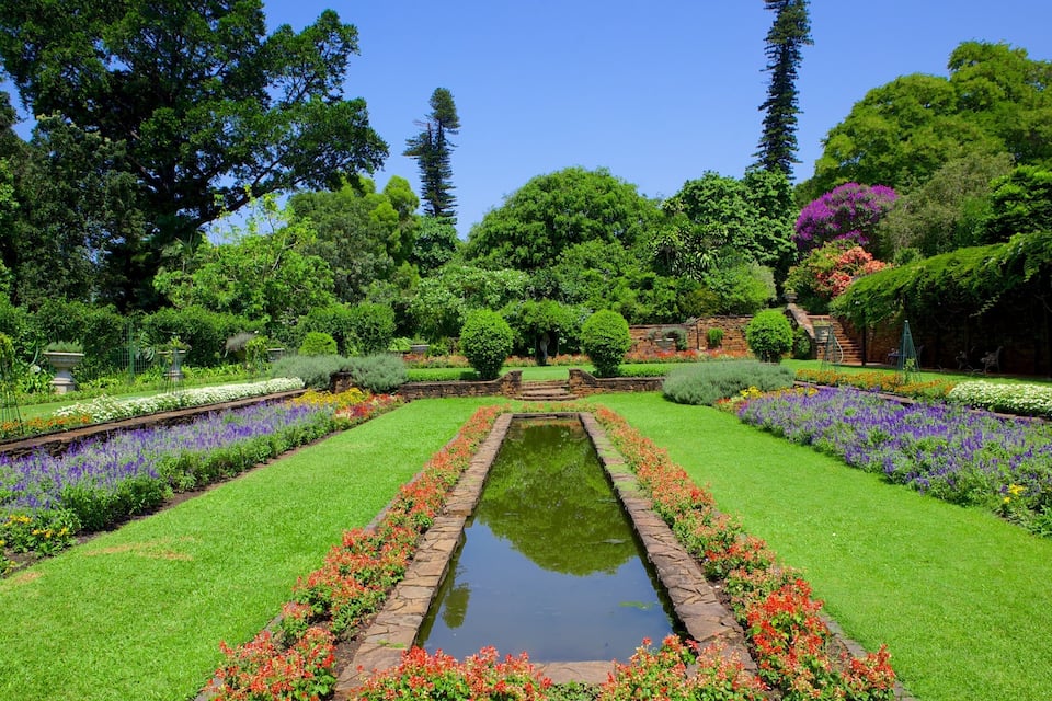 Um jardim formal com um elemento central de água, rodeado por relvados bem cuidados e canteiros de flores coloridos.